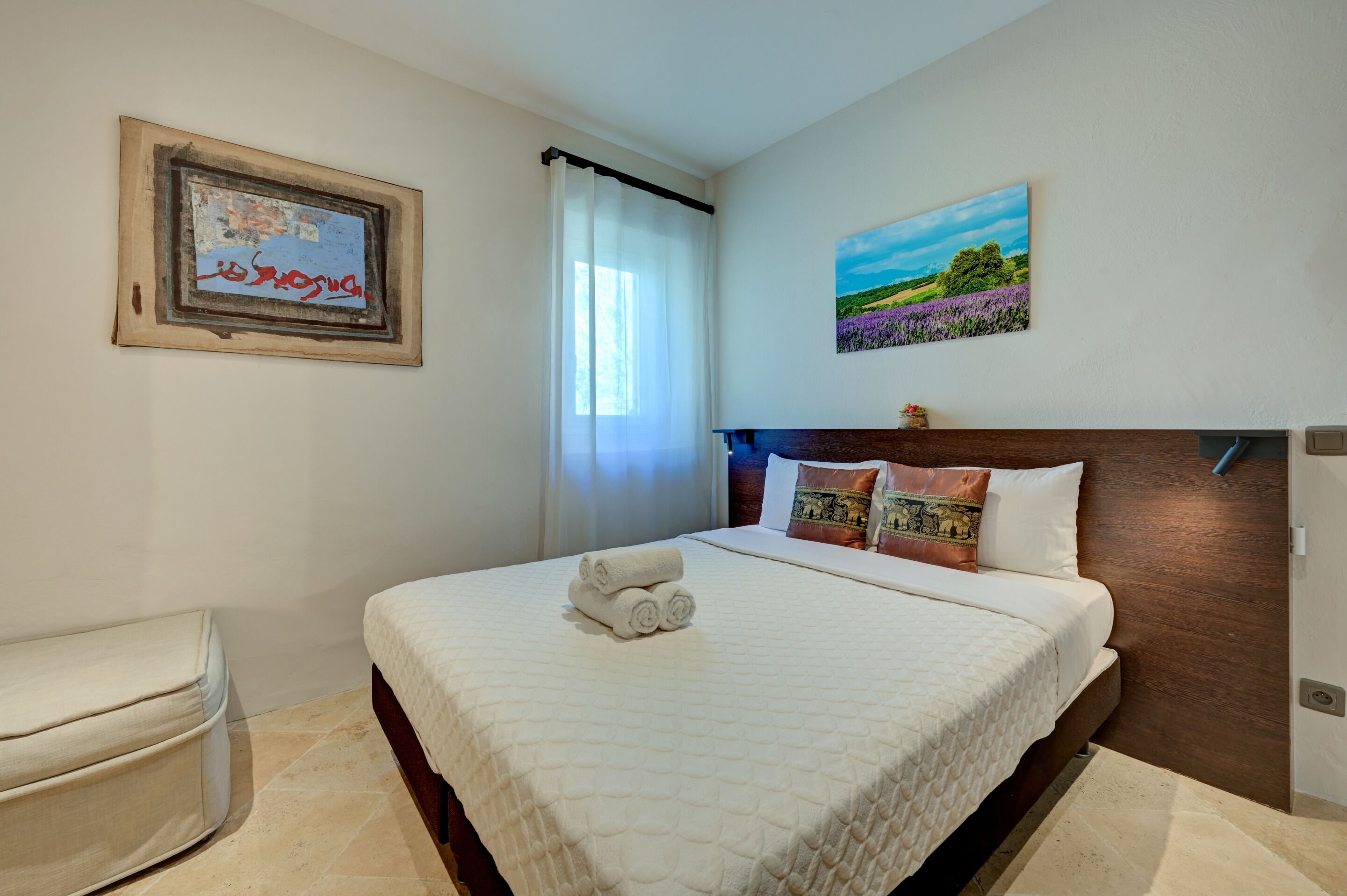 Uma cama bem arrumada com colcha branca, cabeceira com almofadas decorativas, um quadro na parede e uma janela com cortinas leves.