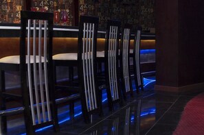 Fileira de cadeiras estilo bar com fitas de luz verticais, um balcão e uma parede estampada ao fundo.