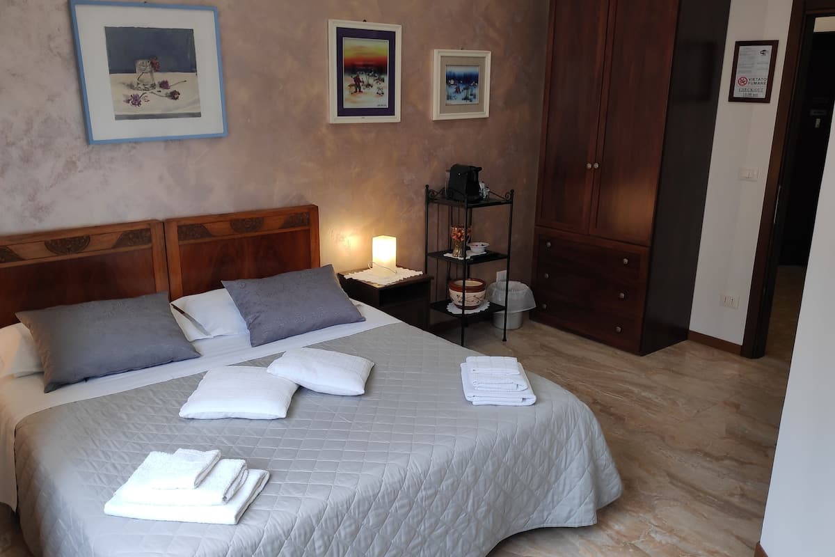 Cama bem arrumada com almofadas e toalhas dobradas, mesa de cabeceira com abajur e uma pequena estante com objetos decorativos.