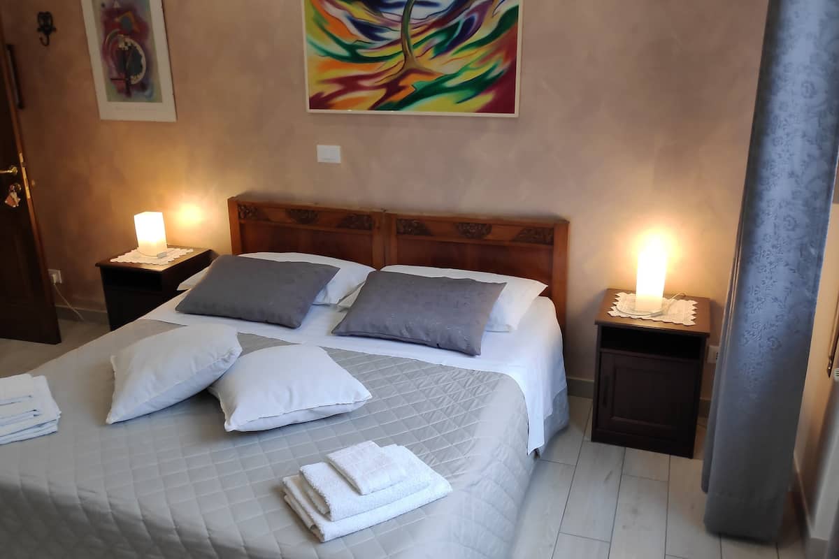 Cama bem arrumada com almofadas e toalhas dobradas, duas luminárias de cabeceira, uma cabeceira de madeira e um quadro abstrato colorido na parede.