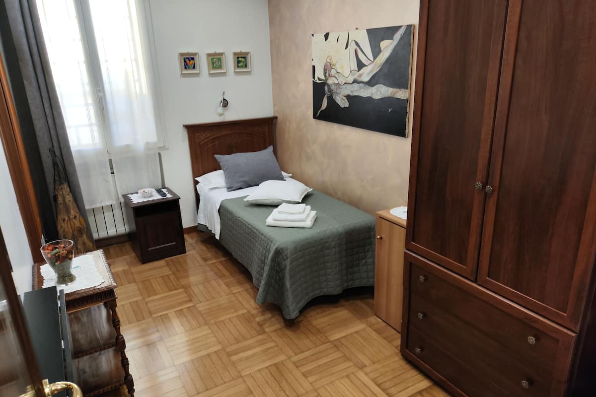 Um quarto com piso de madeira, uma cama com colcha verde, uma mesa de cabeceira com abajur e um guarda-roupa.