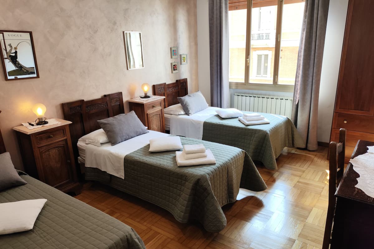 Quarto de hotel com duas camas, piso de madeira, janela com cortinas, mesa de cabeceira com abajur e um quadro na parede.
