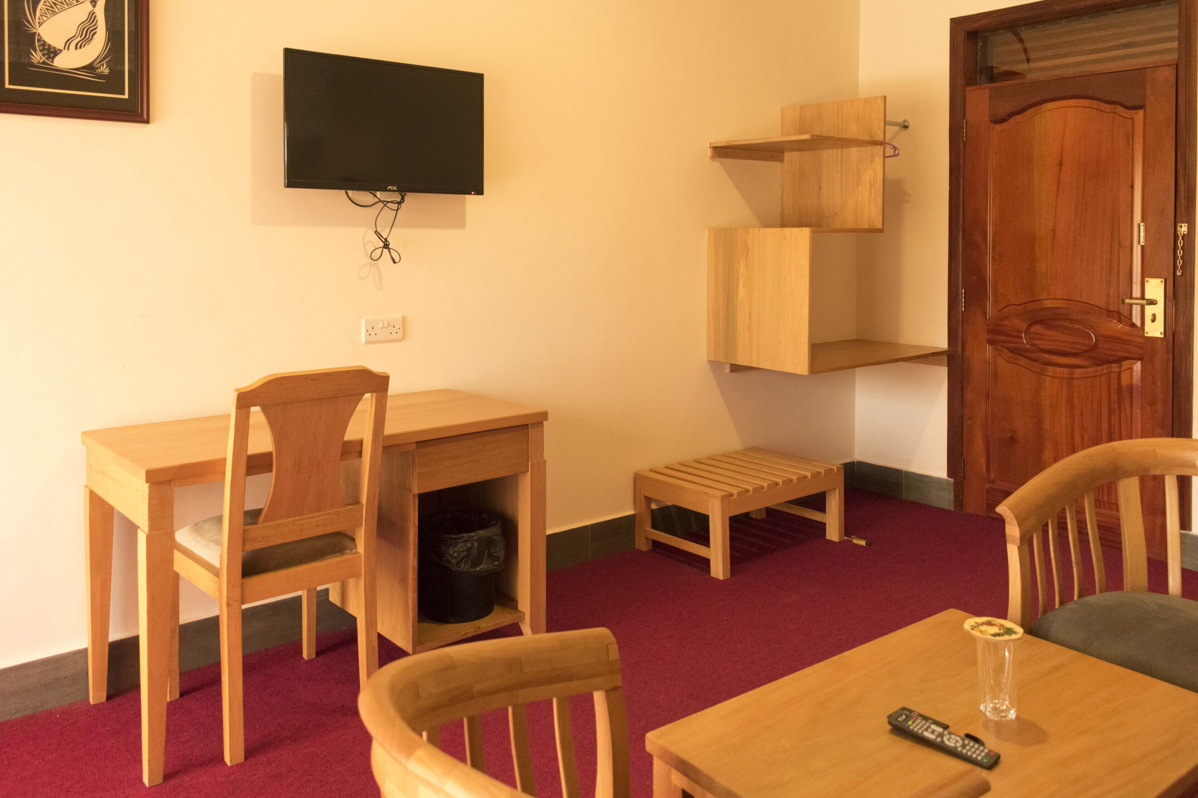 Quarto com escrivaninha de madeira, duas cadeiras, televisão na parede, porta de madeira e um pequeno estante de madeira.