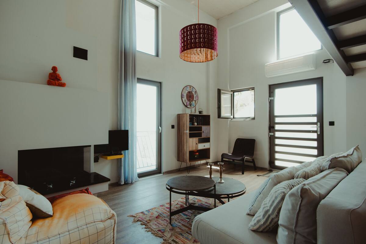 Uma sala de estar moderna com sofá, mesa de centro e televisão. Há uma estante, uma poltrona e uma janela grande.