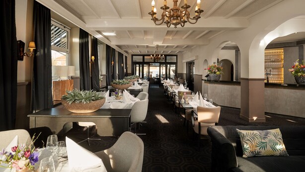 Ampla sala de jantar de hotel, com várias mesas preparadas para refeições, um candelabro e vista da rua através de grandes janelas.