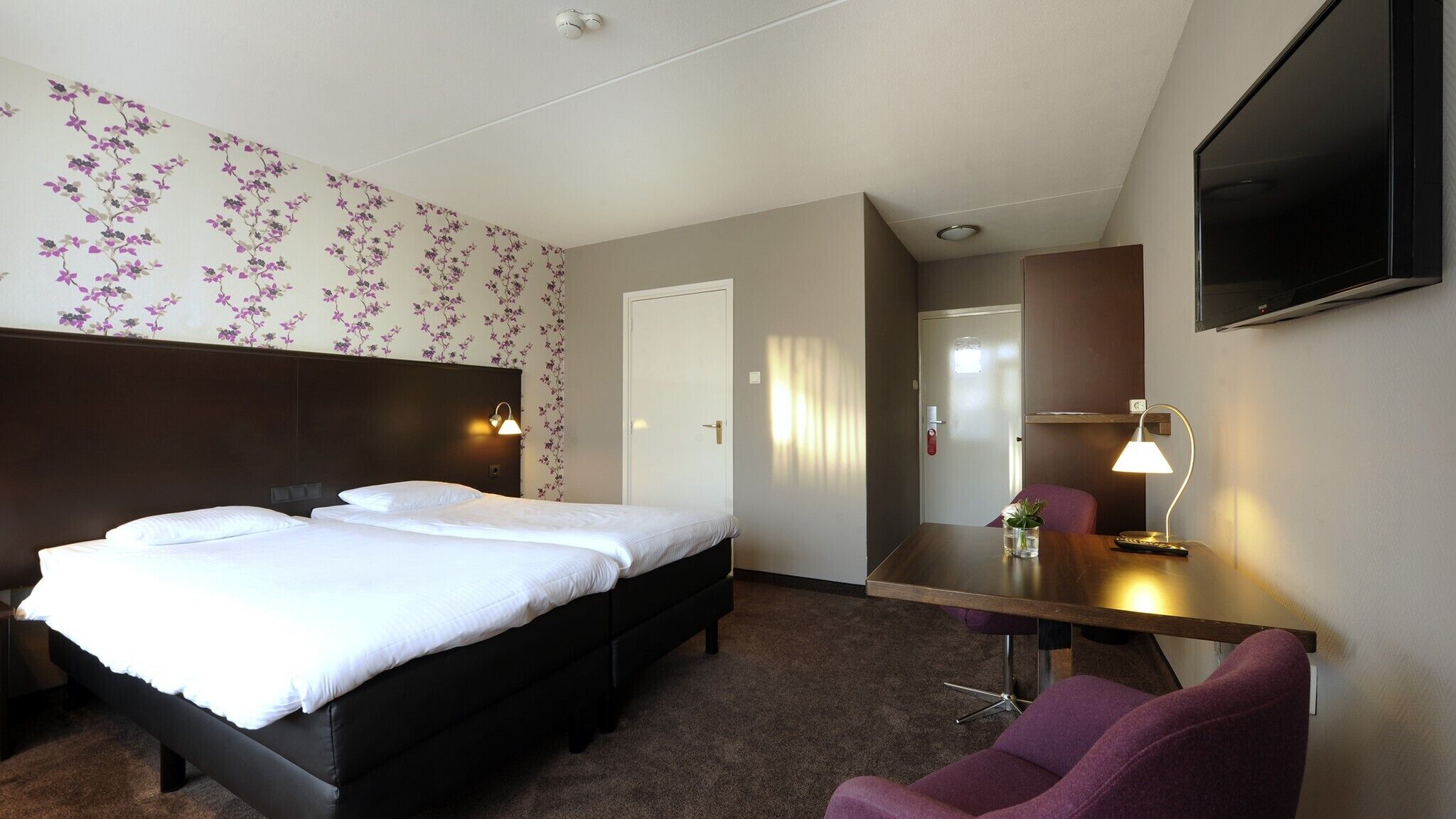 Quarto de hotel com cama de casal, escrivaninha com luminária, cadeira e televisão fixada na parede.