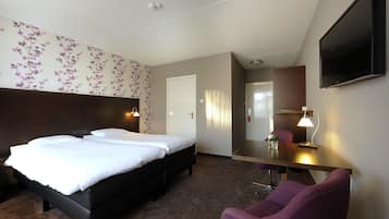 Quarto de hotel com cama de casal, escrivaninha com luminária, cadeira e televisão fixada na parede.