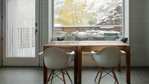 Uma área de refeições com uma mesa de madeira e duas cadeiras brancas, uma porta de vidro deslizante e vista para uma paisagem montanhosa coberta de neve.
