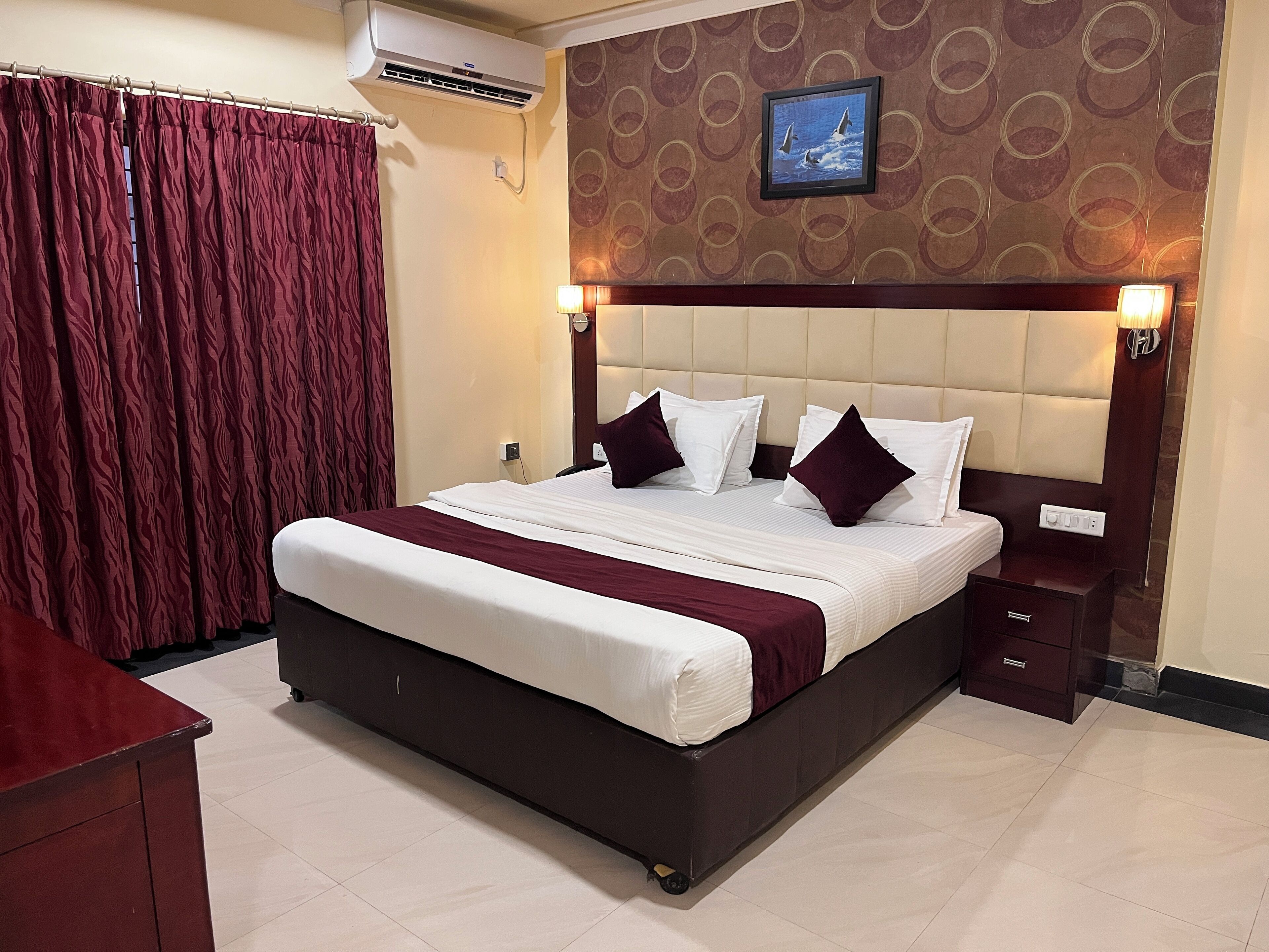 Quarto de hotel com uma cama grande, uma mesinha de cabeceira, uma escrivaninha, uma cadeira, uma cortina vermelha, um aparelho de ar condicionado e uma parede com padrão circular.