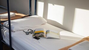 Uma cama de solteiro com colchão branco, manta marrom, um par de óculos, um smartphone e uma revista aberta.