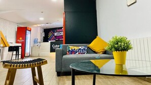 Um hall de hotel moderno com um sofá, uma mesinha e uma planta decorativa. É visível um pilar vermelho e uma placa com a inscrição 'Welcome'.