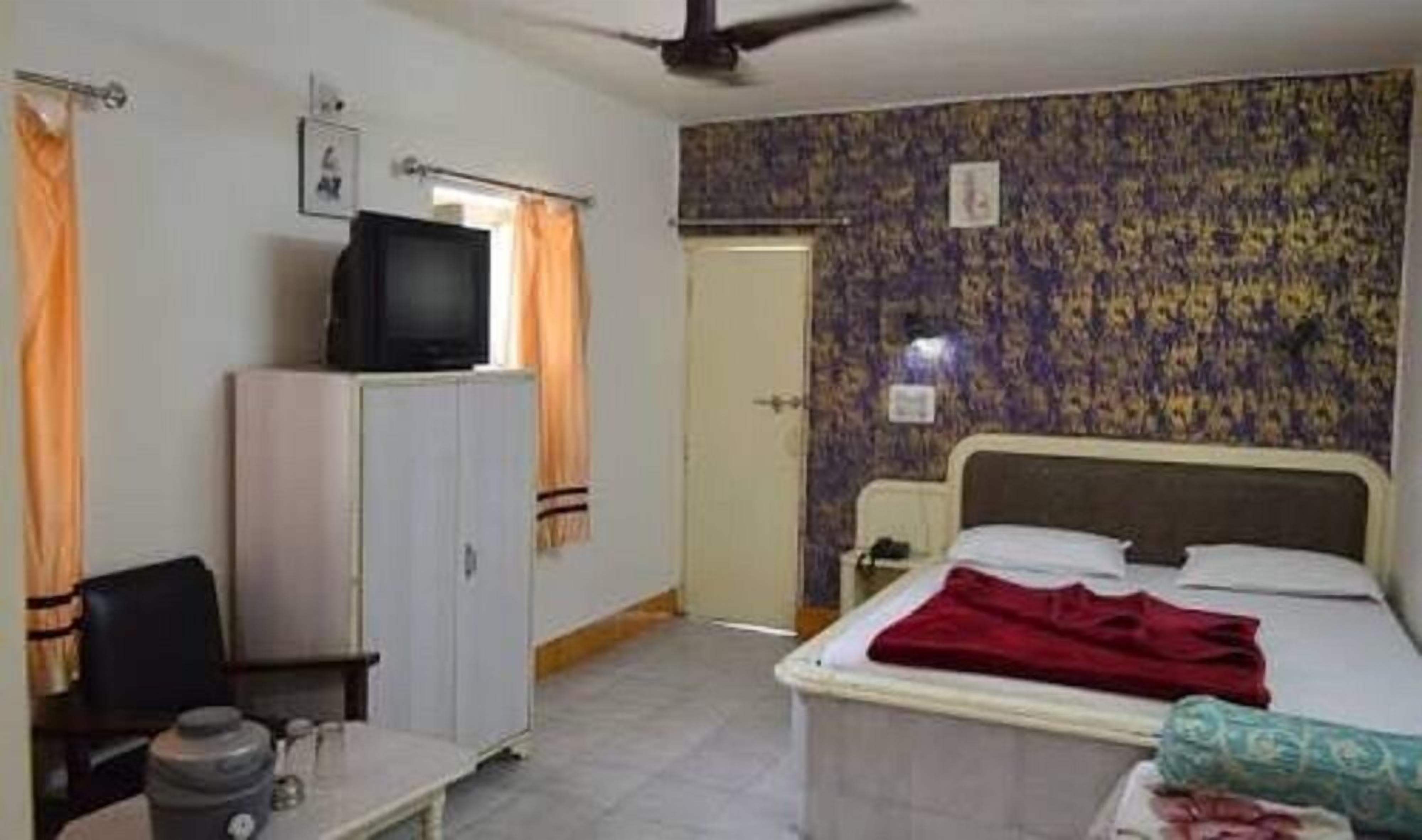 Quarto pequeno e simples com uma cama, uma escrivaninha, uma cadeira, televisão sobre um móvel e uma porta que dá para outro cômodo.