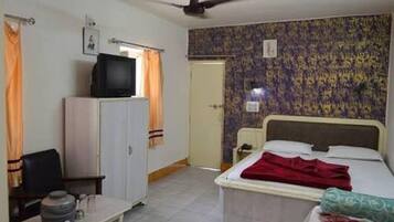Quarto pequeno e simples com uma cama, uma escrivaninha, uma cadeira, televisão sobre um móvel e uma porta que dá para outro cômodo.