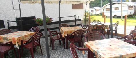Uma área de estar externa com mesas e cadeiras, coberta por uma cobertura, e trailers de recreação ao fundo.
