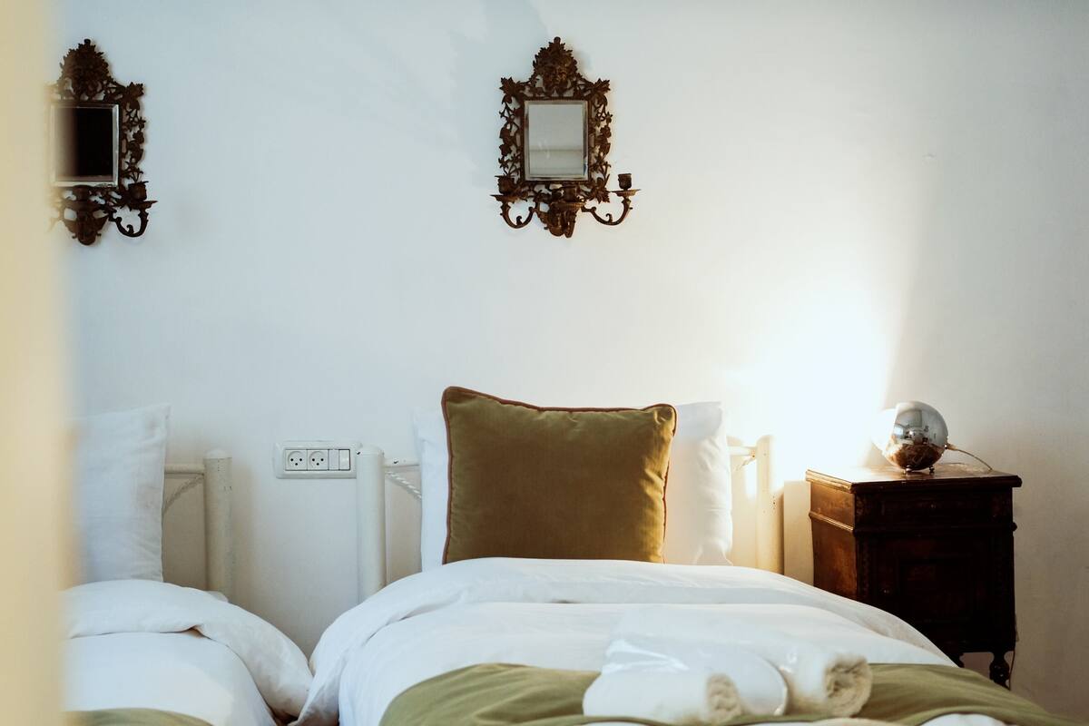 Quarto com duas camas, uma mesinha de cabeceira com abajur e um espelho fixado na parede.