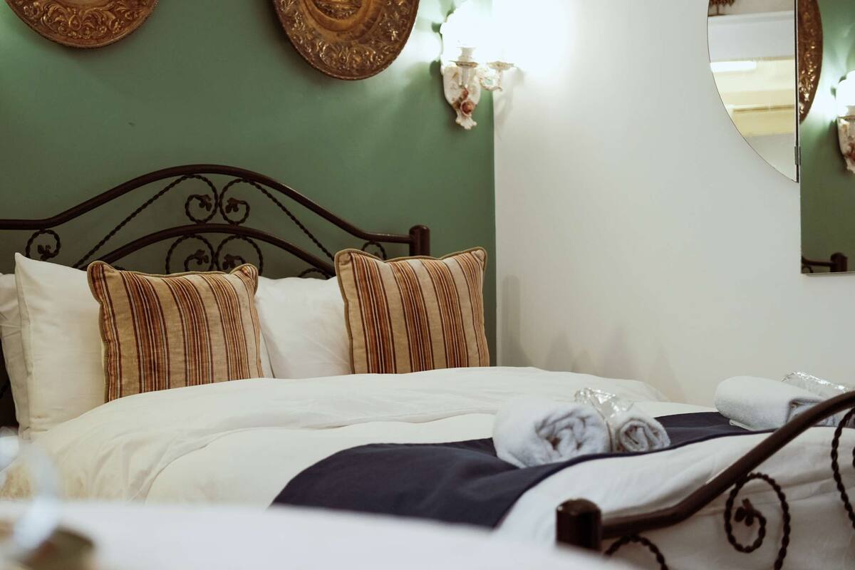 Cama bem arrumada com almofadas listradas, cabeceira escura e parede verde com arandelas decorativas.