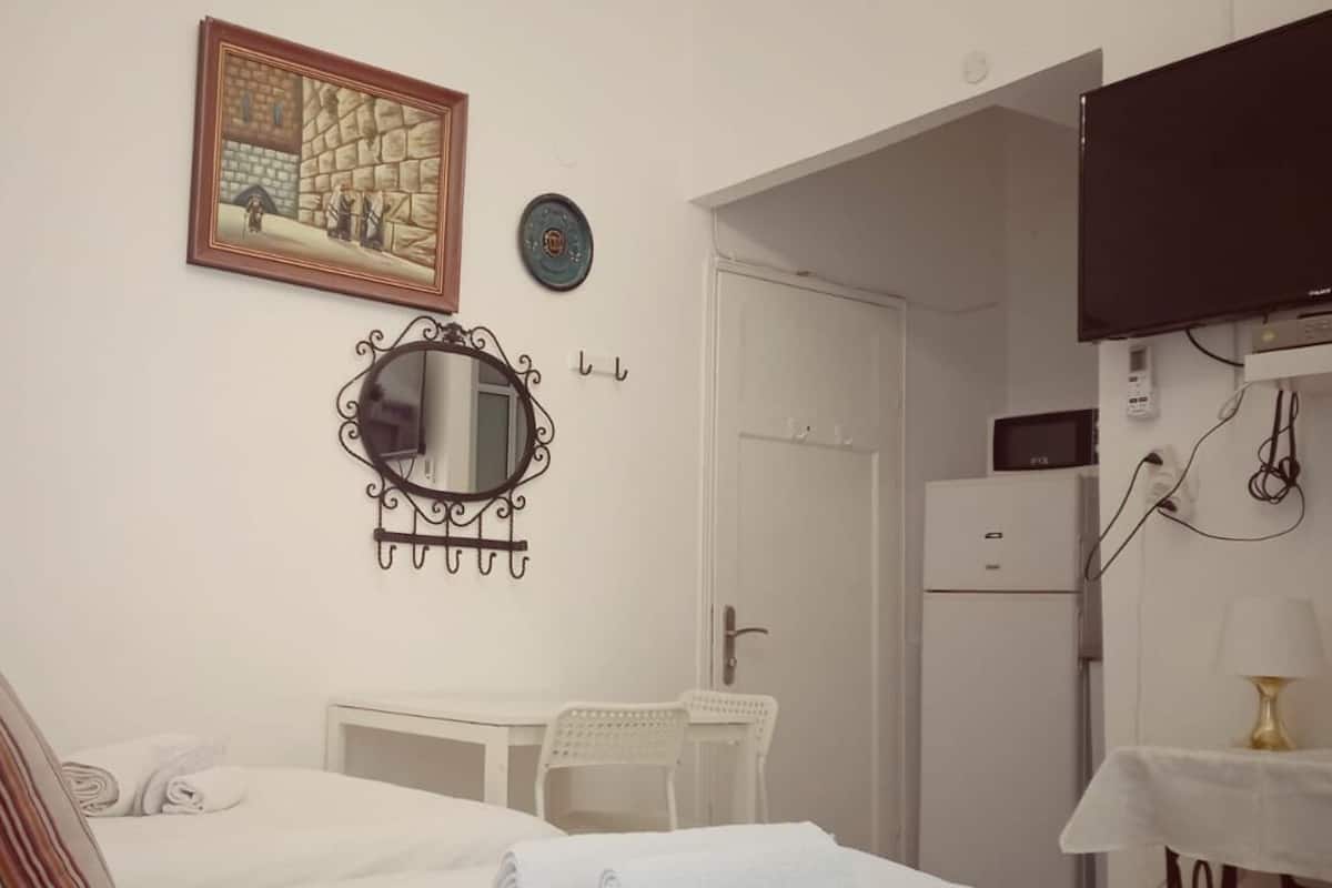 Quarto de hotel com cama, uma pequena mesa, cadeira, espelho e porta para uma área de cozinha.