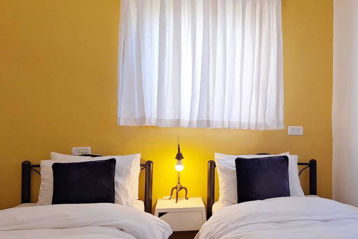 Uma cama de casal com roupa de cama branca, duas mesas de cabeceira, um abajur e uma janela com cortinas brancas.
