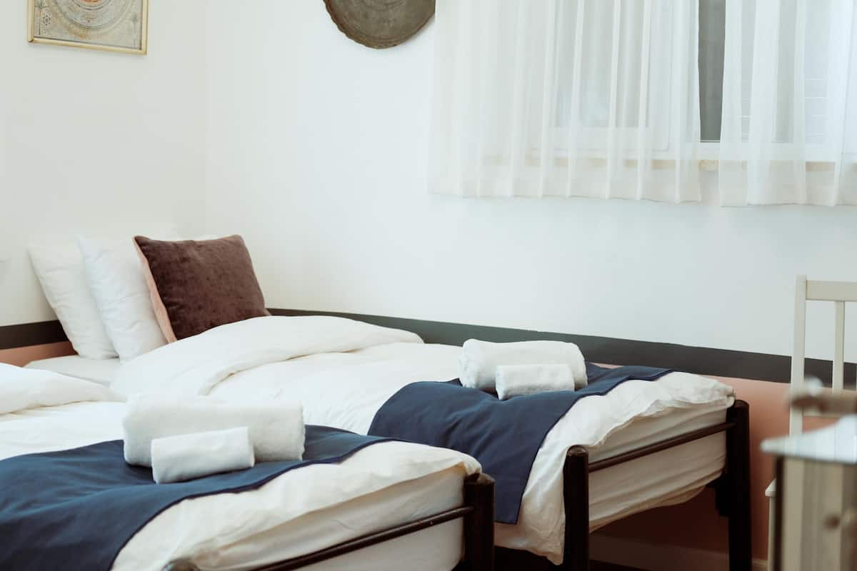 Quarto de hotel com duas camas, roupa de cama branca, travesseiro marrom, manta azul, cabeceira de madeira, uma mesinha, uma cadeira e uma janela com cortinas translúcidas.