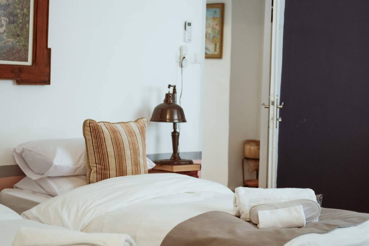 Cama bem arrumada com roupa de cama branca e bege, um travesseiro listrado, uma luminária de mesa em latão e uma pintura na parede.