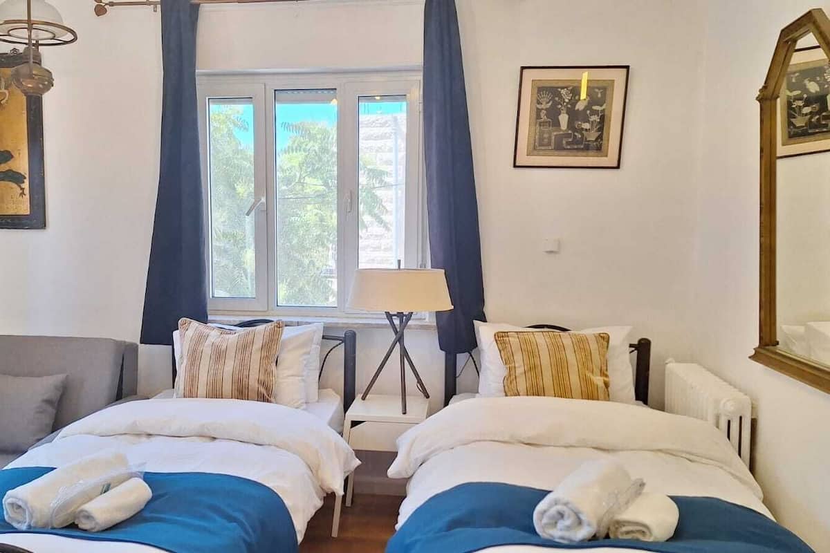Quarto de hotel com duas camas, um sofá, uma janela com cortinas, um abajur e obras de arte nas paredes.
