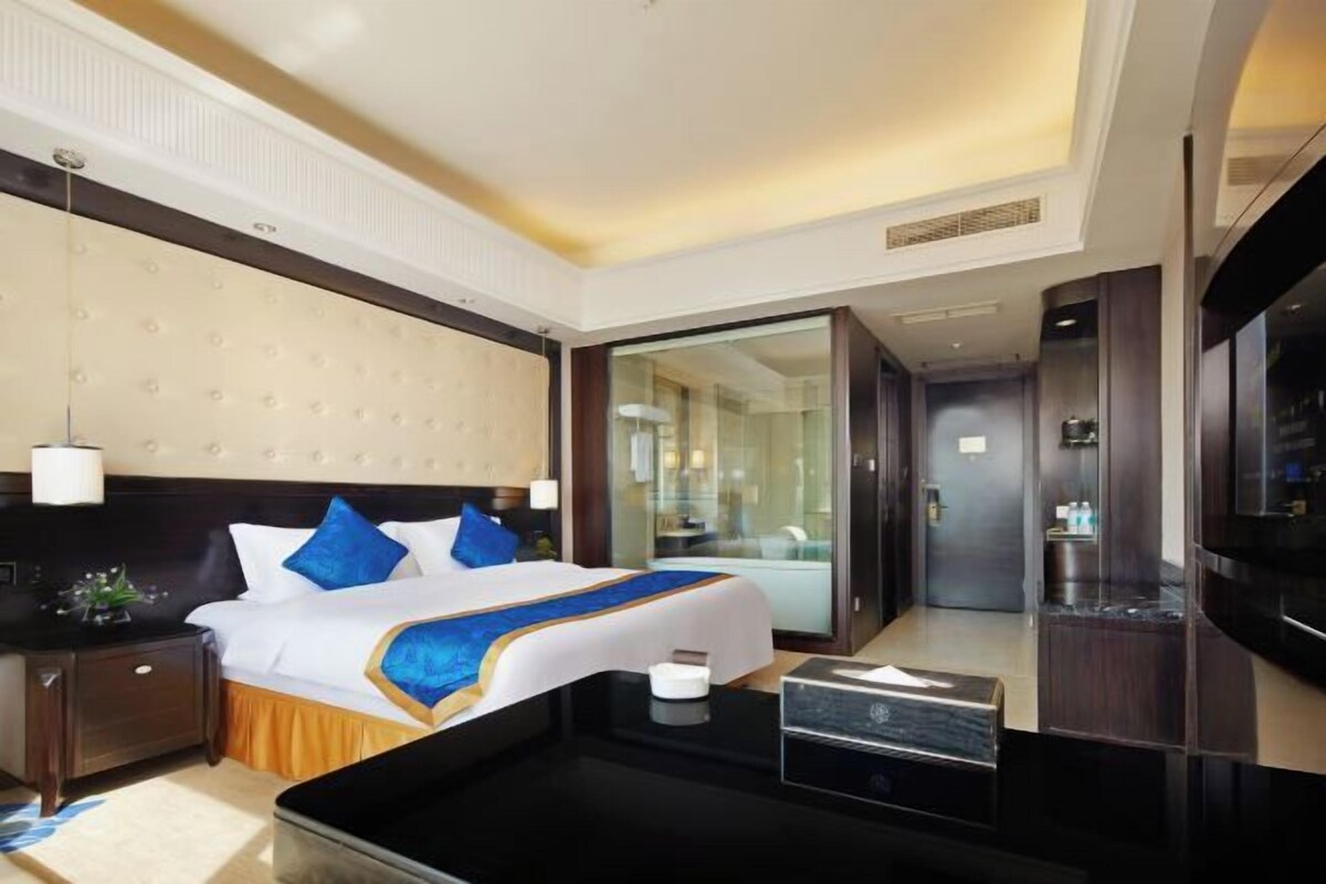 Quarto de hotel com uma cama grande, televisão, mesa de cabeceira e banheiro com paredes de vidro.