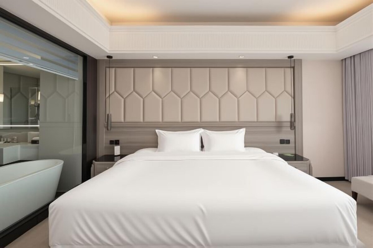 Quarto de hotel moderno com uma cama grande, uma cabeceira embutida, uma mesa de cabeceira e um banheiro com banheira.