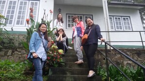 Um grupo de pessoas em uma escadaria, com uma parede de pedra e um edifício com janelas brancas ao fundo.