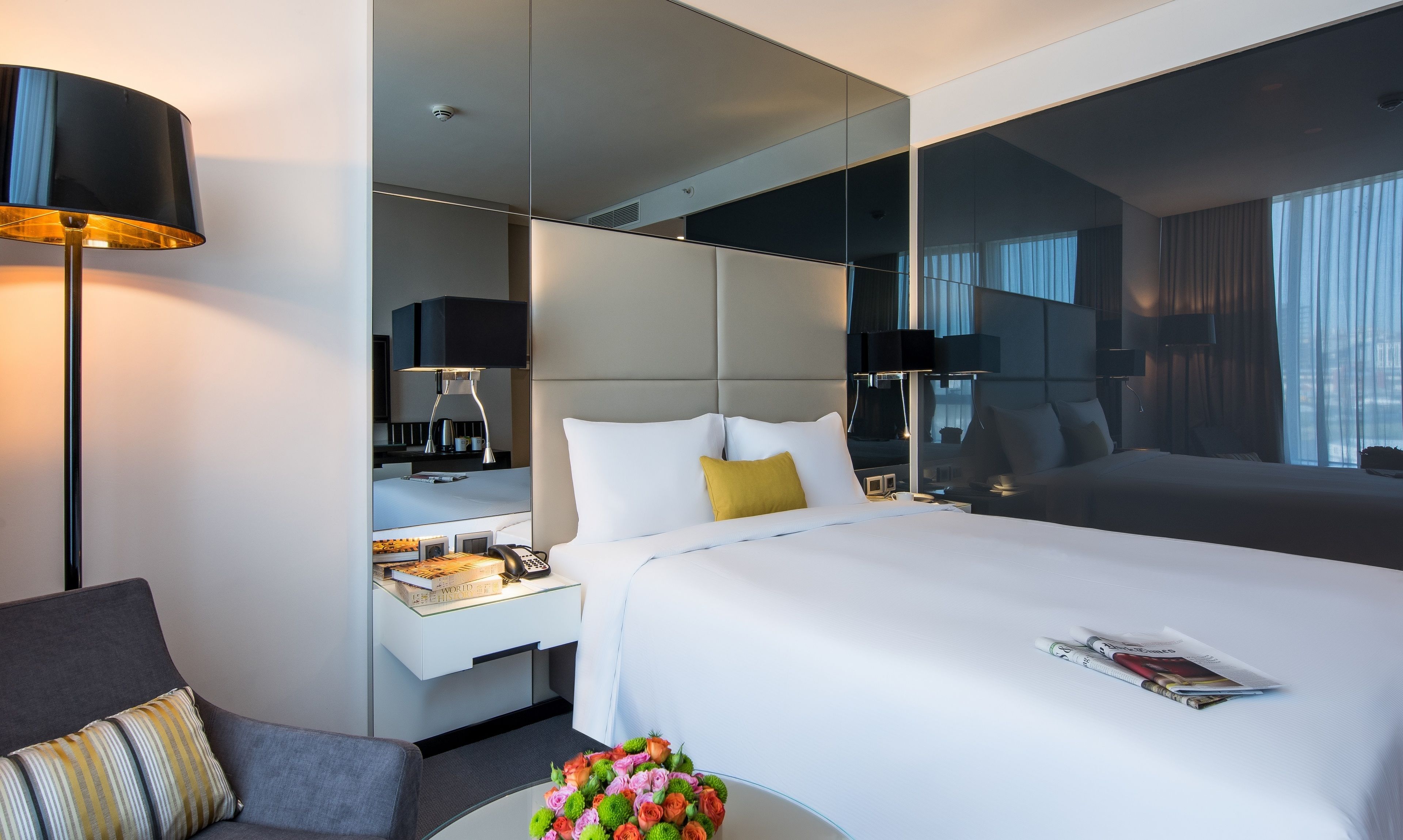 Quarto de hotel moderno com uma cama grande, uma mesa de cabeceira com abajur, uma escrivaninha com computador e um espelho amplo.