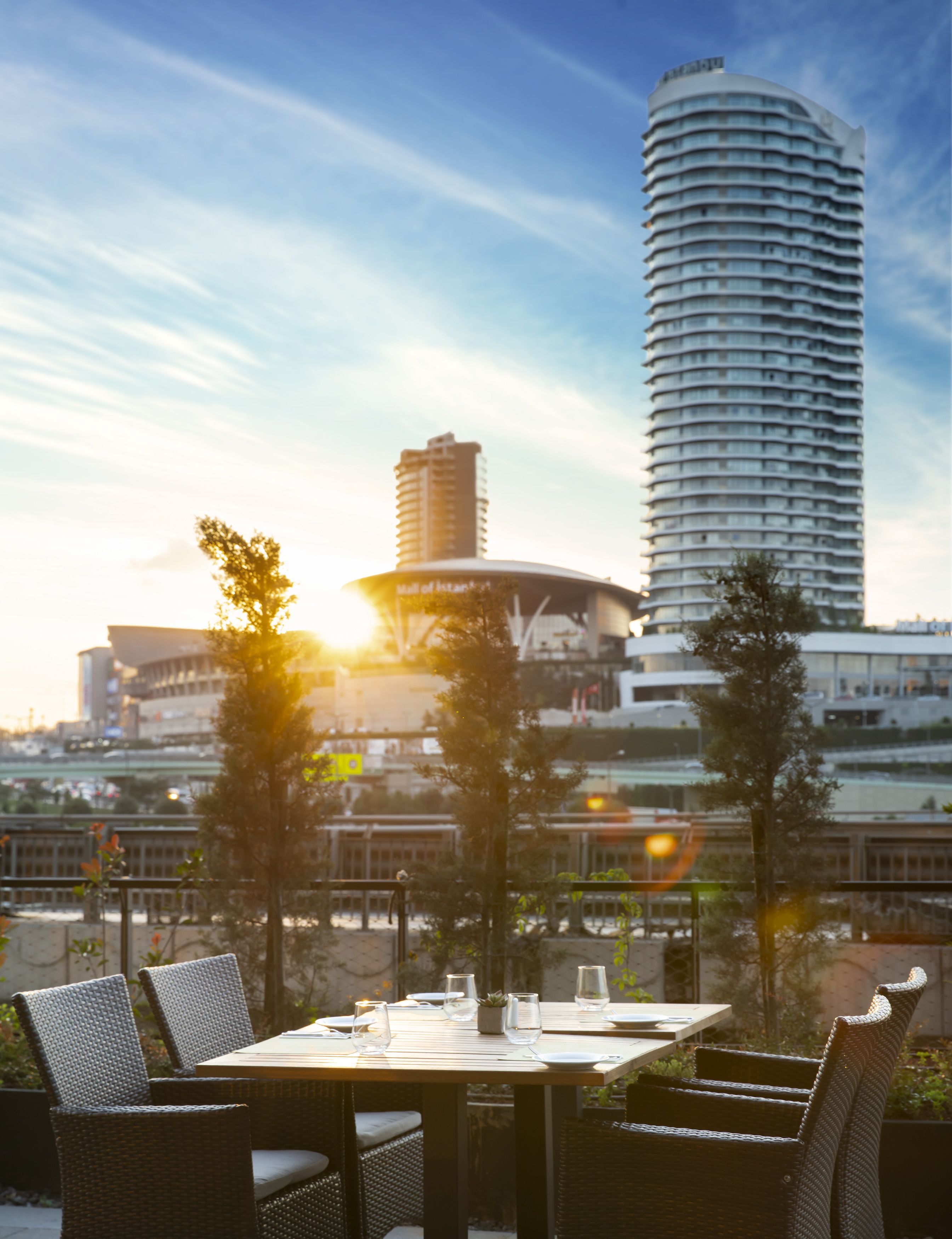 Uma área de refeições ao ar livre com cadeiras de vime e uma mesa para dois, com vista para um horizonte urbano repleto de edifícios modernos e céu claro.