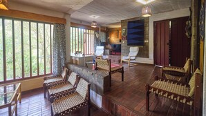 Quarto com piso de madeira, várias cadeiras de vime, um sofá, uma mesa de jantar e uma grande janela com vista para o verde.