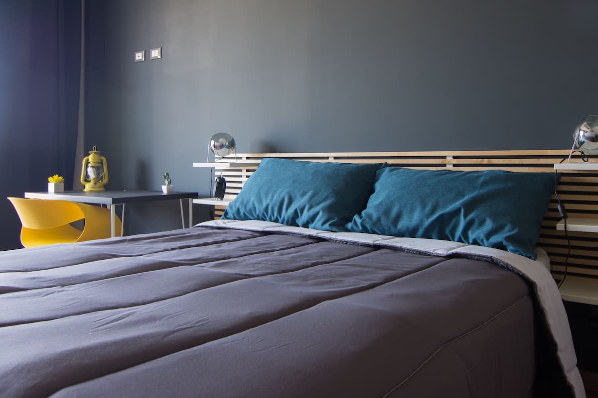 Quarto de hotel moderno com uma cama grande, uma mesa de cabeceira, uma luminária e uma pequena mesa com uma cadeira amarela.