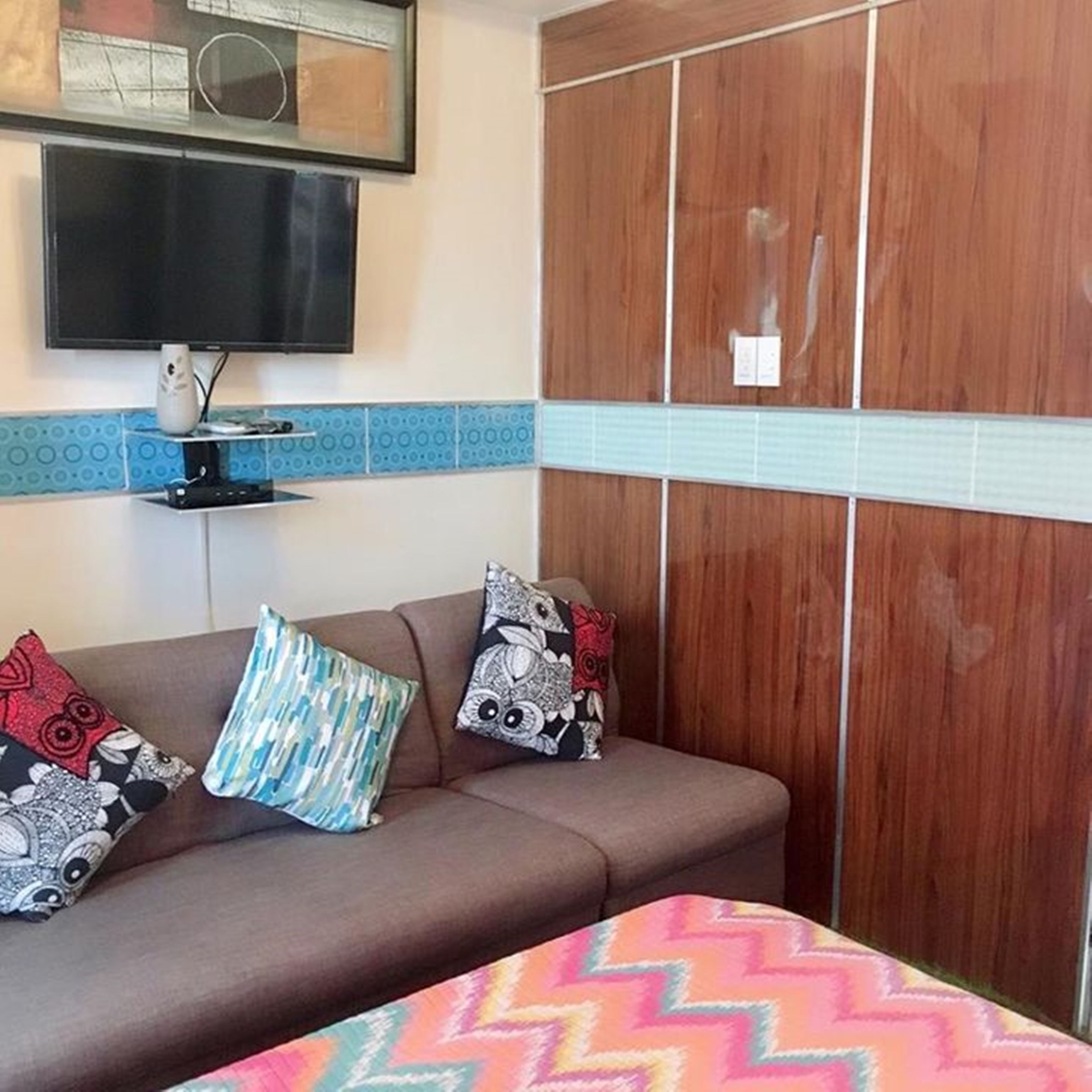 Quarto com sofá marrom, almofadas estampadas, televisão na parede e guarda-roupa de madeira.
