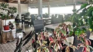 Uma área de refeições interna com mesas, cadeiras, plantas em vasos e um teto alto com vigas aparentes.