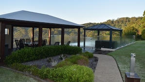 Uma área externa coberta com uma mesa e cadeiras, um gazebo com paredes de vidro e vista para uma colina arborizada.