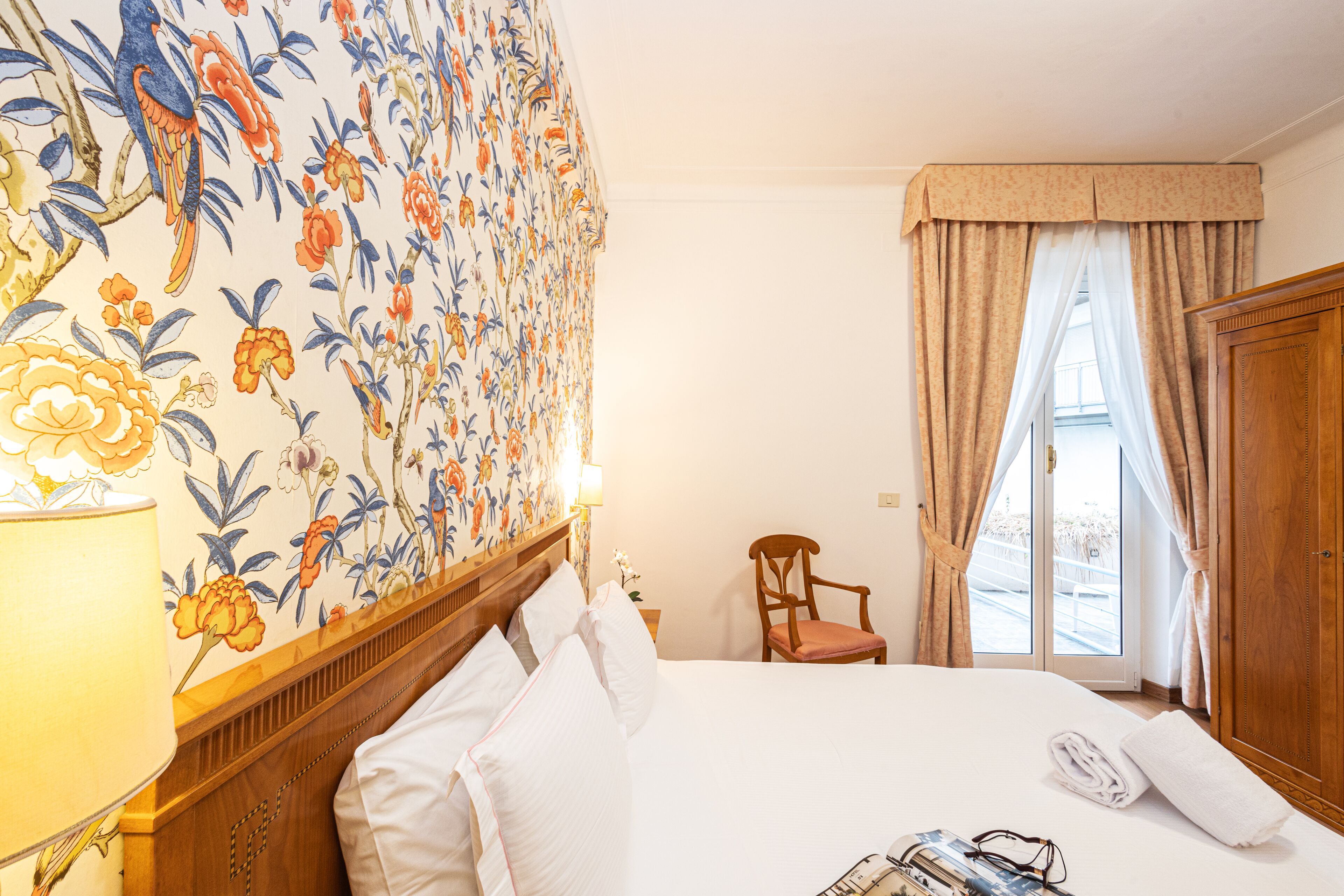 Quarto de hotel com papel de parede floral, cabeceira de madeira, cama com roupa de cama branca, cadeira de madeira, janela com cortinas e guarda-roupa de madeira.