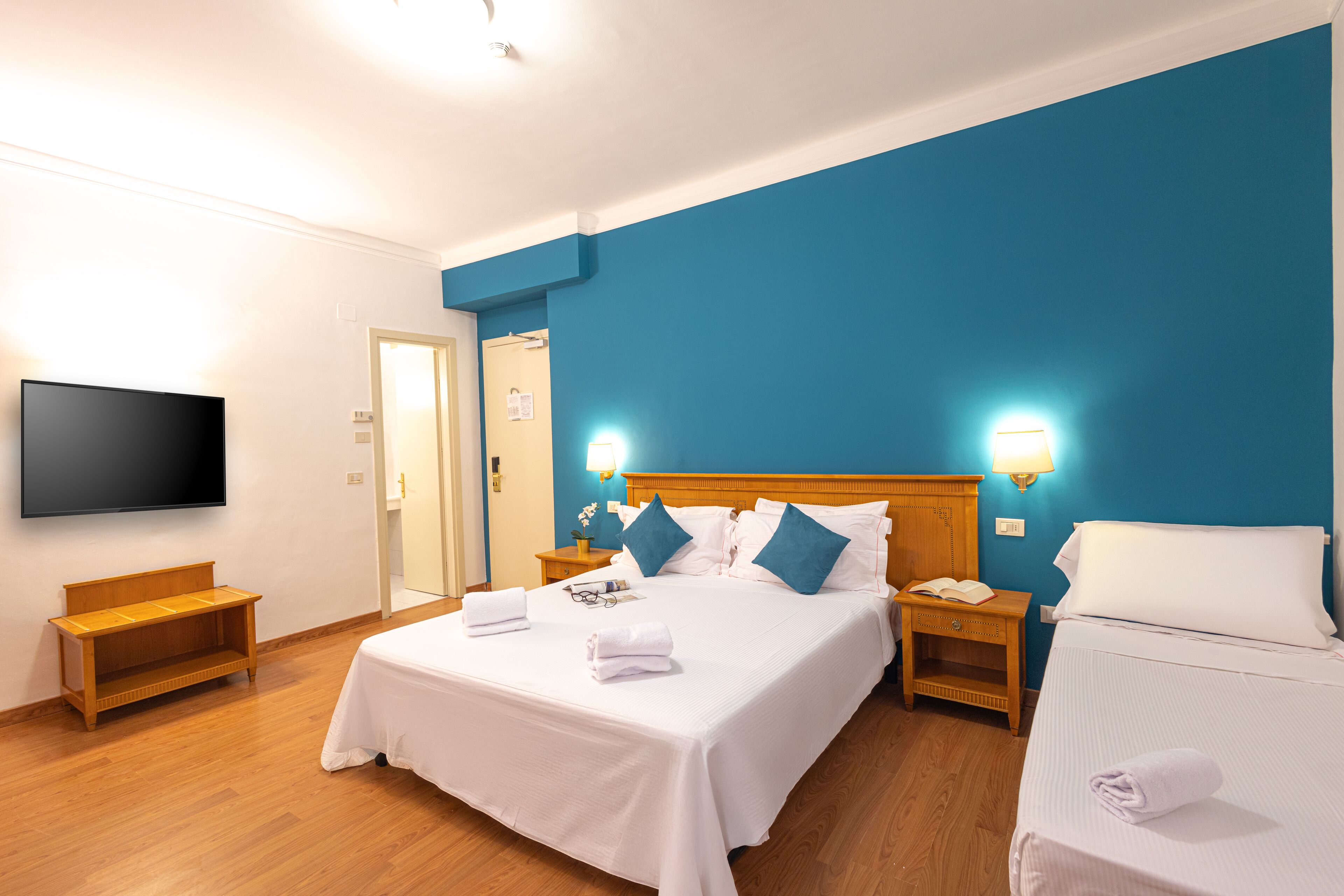 Quarto de hotel com uma cama grande, mesas de cabeceira, televisão de tela plana e um banco de madeira.