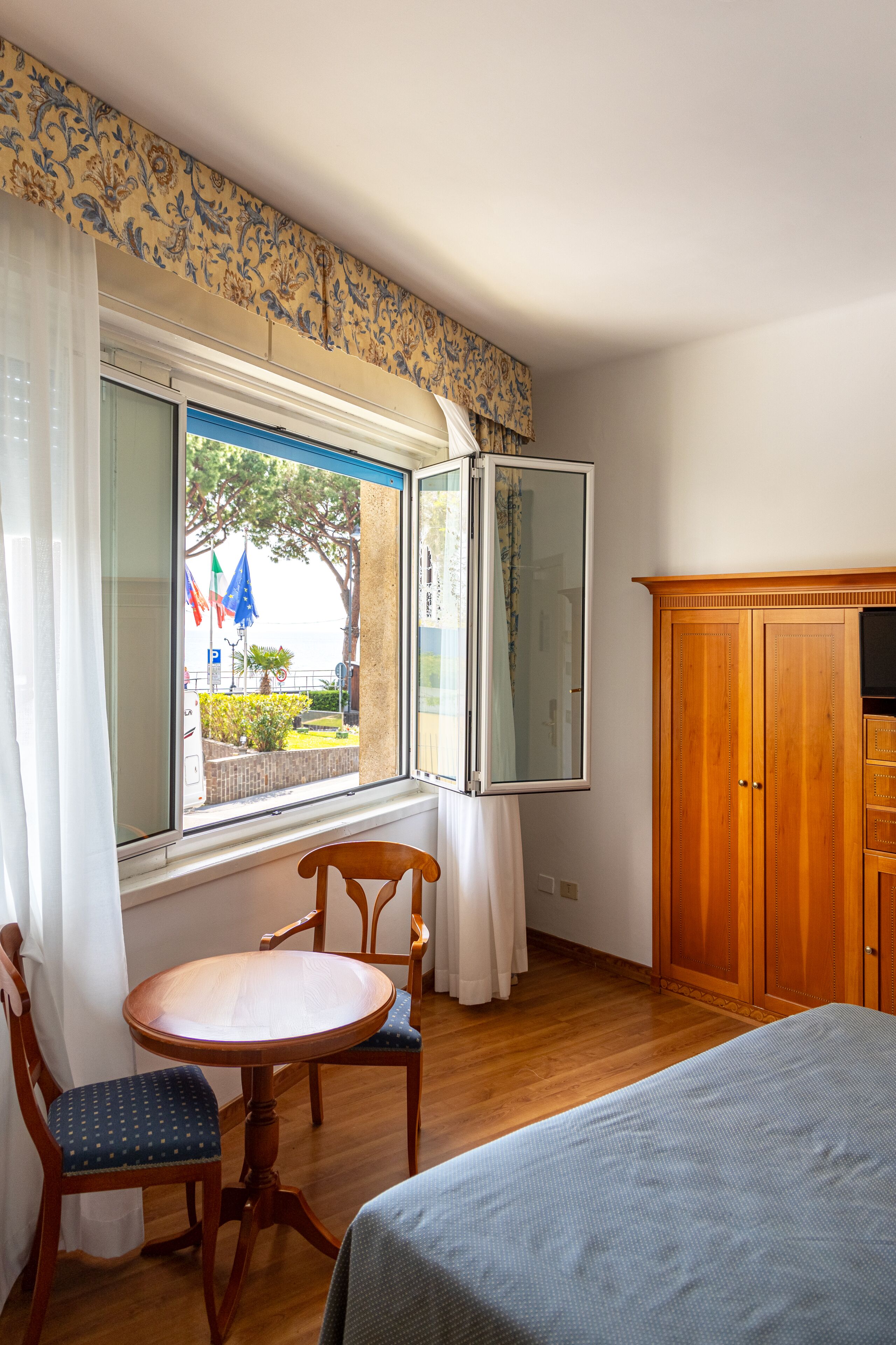 Um quarto com um armário de madeira, uma cama, uma mesa redonda com cadeiras e uma janela com cortinas.
