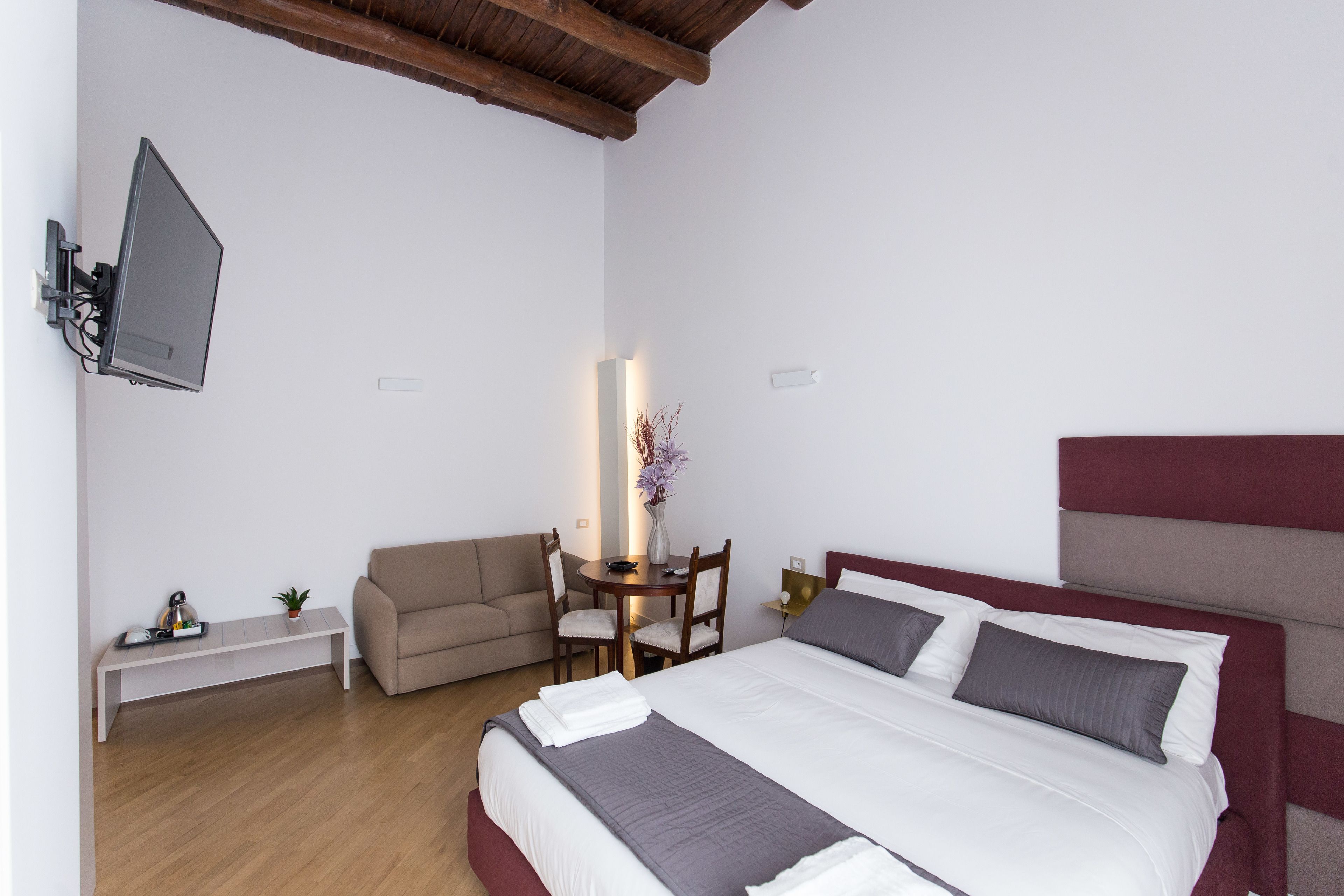 Quarto de hotel com cama, sofá, escrivaninha com cadeira, TV na parede e um vaso com flores.