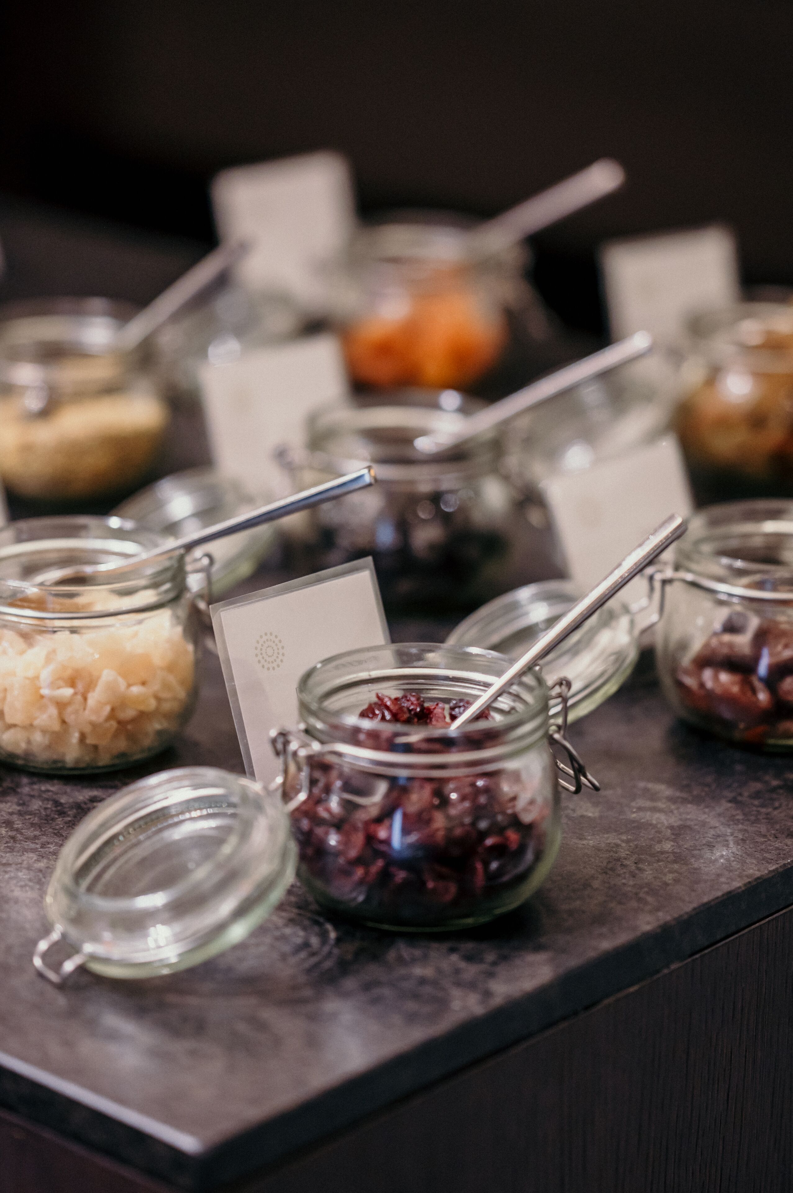 Uma mesa de buffet com diversos alimentos em recipientes de vidro, cada um com etiqueta e uma colher.
