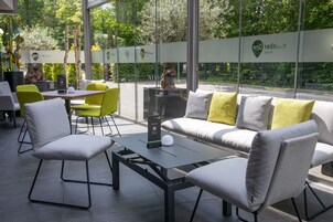 Uma área de estar moderna ao ar livre, com sofá, cadeiras e mesas, exibindo um sinal com código QR e vista para o verde ao redor.