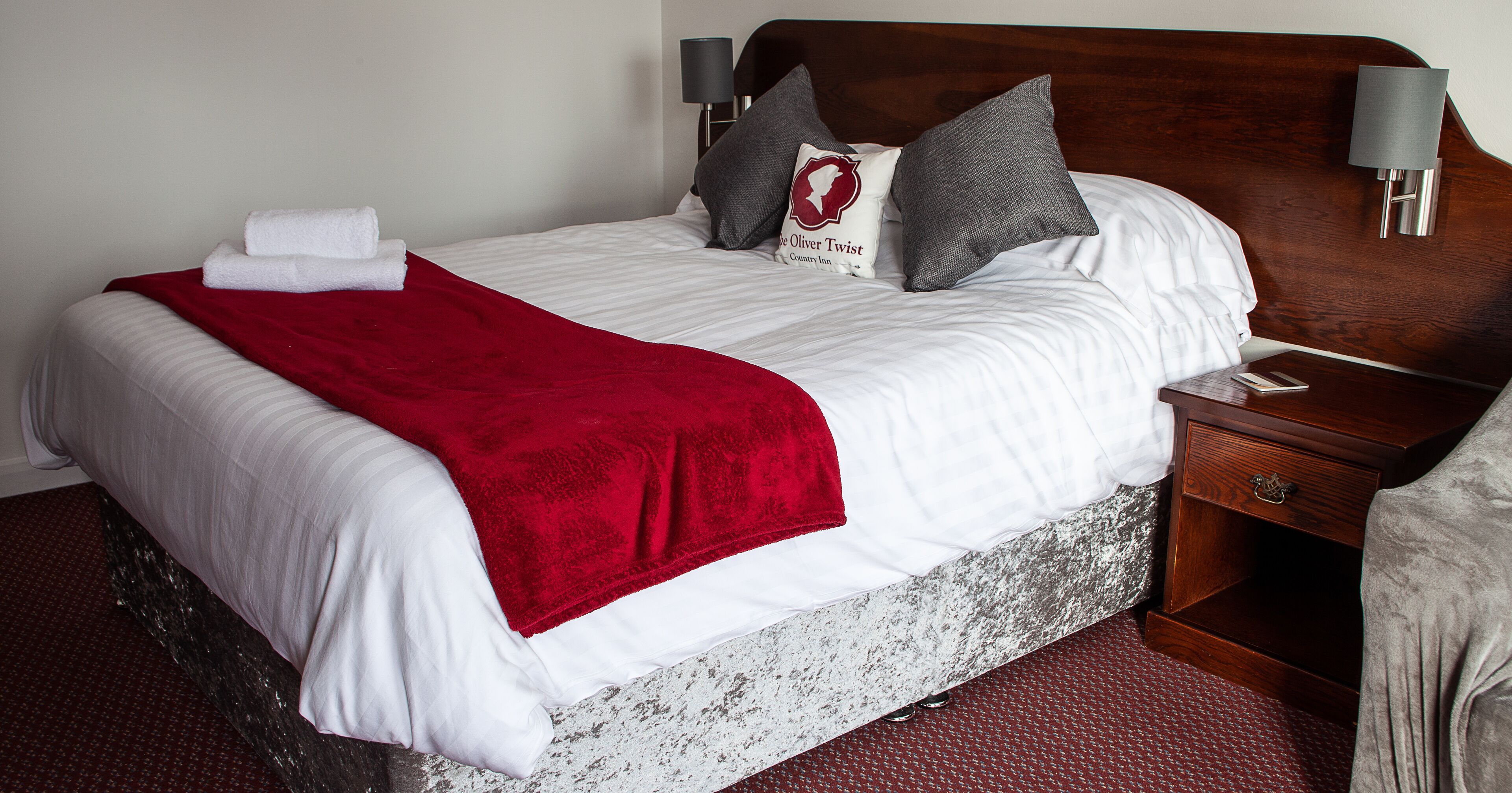 Cama bem arrumada com lençóis brancos, manta vermelha e duas almofadas, uma delas com logotipo. Há uma mesa de cabeceira de madeira com luminária e gaveta.
