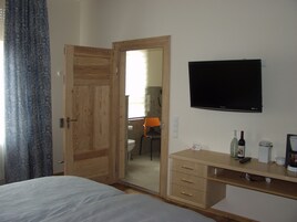 Quarto com porta de madeira, uma cama, TV fixada na parede, escrivaninha com uma garrafa de vinho e uma cadeira ao fundo.
