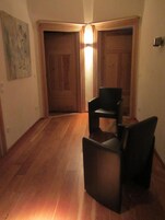 Um corredor com piso de madeira, uma cadeira preta e uma porta com luminária.