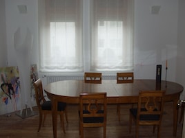 Uma sala de jantar com uma mesa redonda, seis cadeiras de madeira, duas janelas com cortinas finas e um radiador sob as janelas.