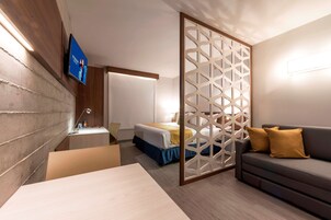 Quarto de hotel moderno com cama, sofá, televisão fixada na parede e um divisória decorativa.