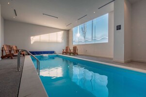 Piscina interior com paredes de vidro transparente, cadeiras de madeira e um ambiente banhado pela luz do sol.