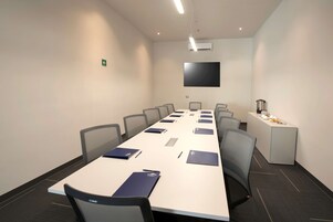 Uma sala de conferências com uma mesa comprida, cadeiras, uma TV de tela plana e um pequeno armário com objetos dentro.