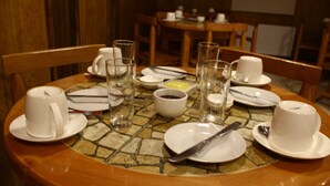 Mesa de jantar posta com pratos brancos, xícaras e talheres, rodeada por cadeiras.