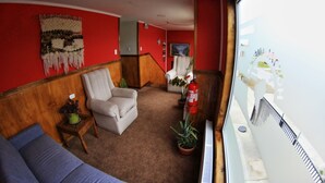Quarto com parede vermelha, sofá cinza, poltrona branca, mesa de madeira pequena com uma planta e um extintor perto de uma janela.